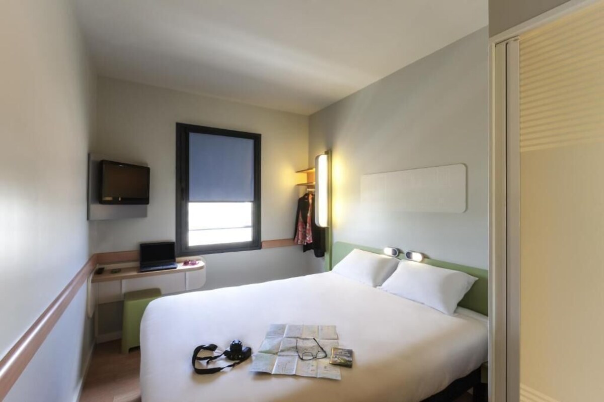 Quarto de hotel com uma cama, uma mesa com um laptop, uma televisão na parede e uma janela que proporciona iluminação natural.
