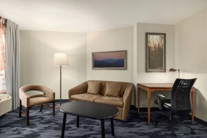 Quarto de hotel com sofá, mesa pequena, escrivaninha, cadeira, luminária e um quadro na parede.