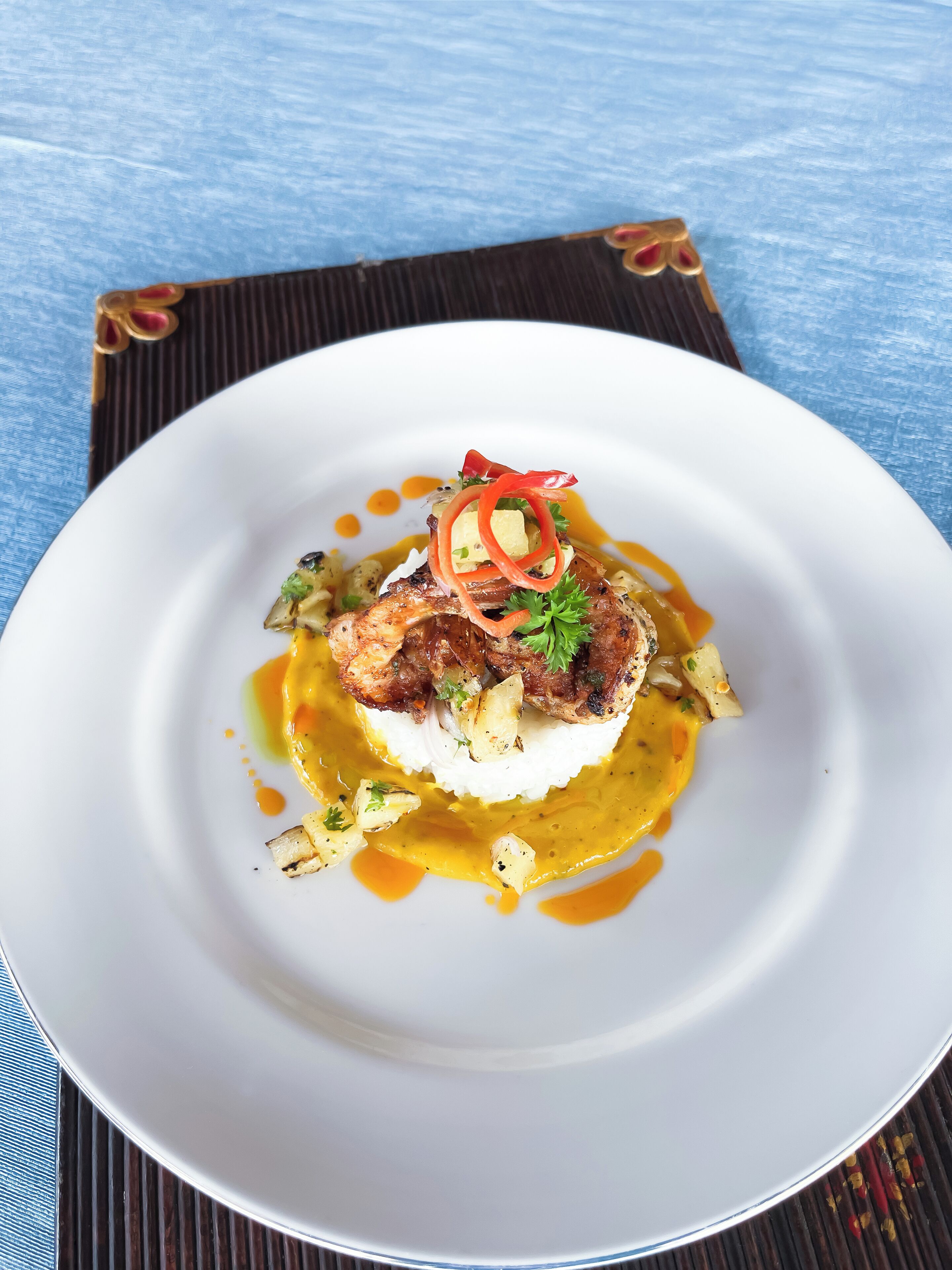 Prato branco com um prato composto por uma base branca, possivelmente arroz, coberto com um pedaço de carne, guarnecido com pimenta vermelha e salsa, e rodeado por um molho amarelo.