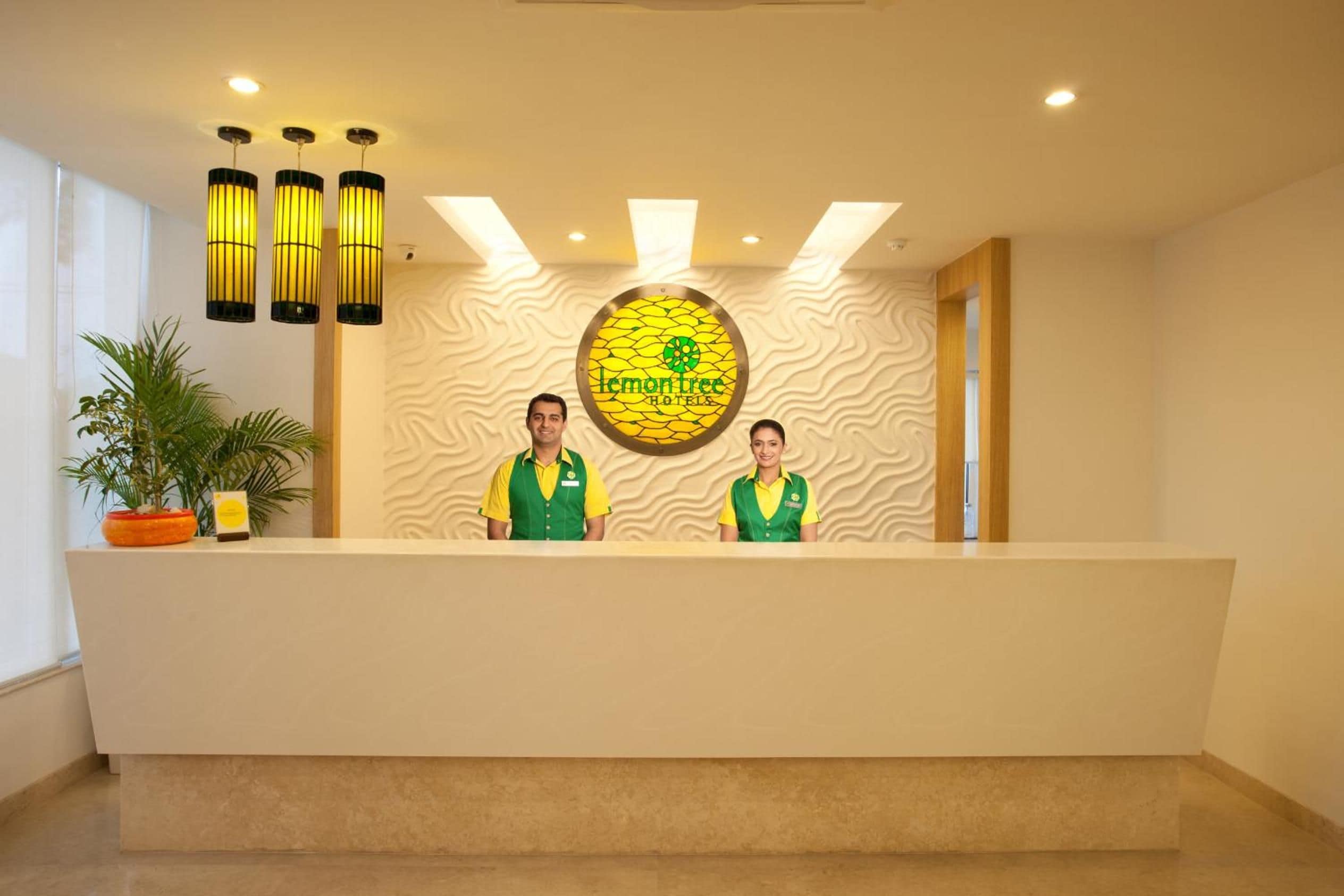 Bancada de recepção com dois funcionários, logotipo na parede e uma planta à esquerda.