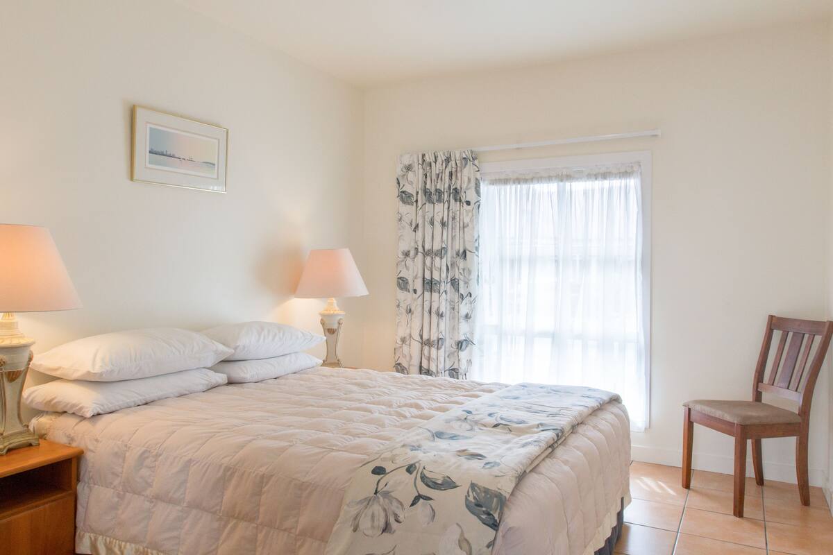 Quarto com cama, duas luminárias de cabeceira, uma cadeira, janela com cortinas e um quadro na parede.