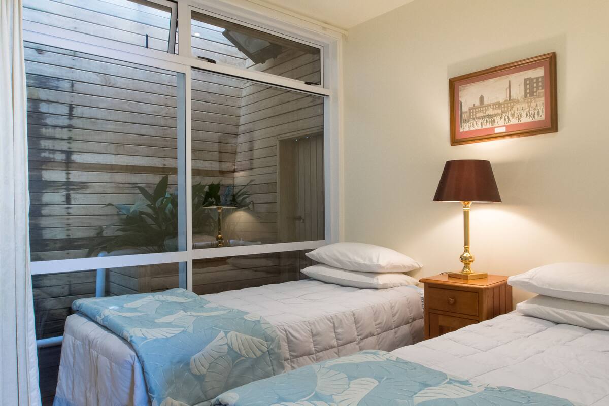 Quarto com duas camas, mesa de cabeceira com abajur, quadro pendurado na parede e janela com persianas.
