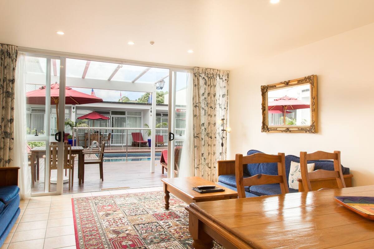 Ampla sala de estar com mesa de jantar de madeira, sofá azul e vista para um pátio externo com guarda-sóis.