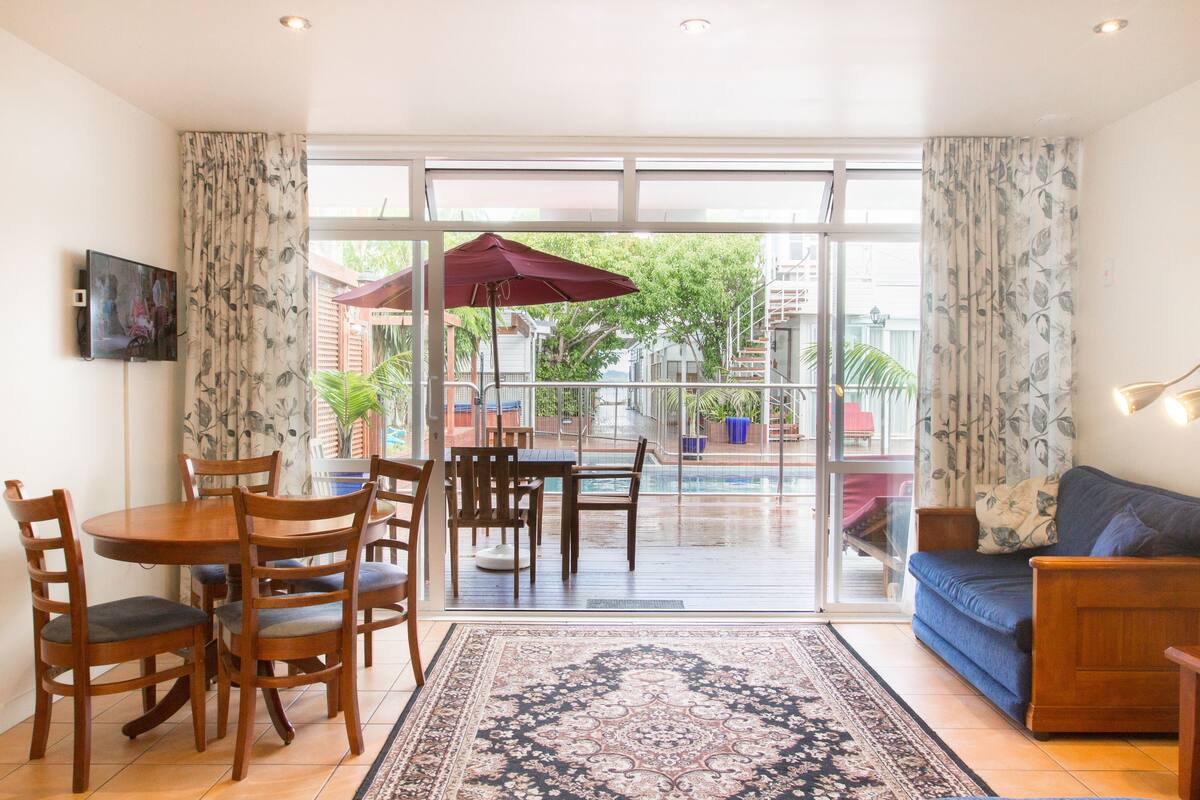 Uma sala de estar com sofá, mesa de jantar e cadeiras, TV fixada na parede e uma grande porta de vidro deslizante que dá para área externa com piscina e guarda-sóis.