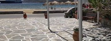 Uma área com piso de pedra, com um banco e plantas em vasos, com vista para um corpo d'água e uma praia.