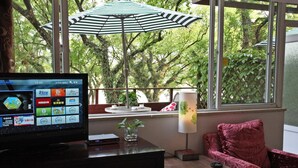 Quarto com televisão exibindo guia de canais, mesa com vaso de vidro, cadeira vermelha e janela com vista para vegetação.