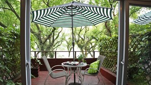 Um pátio externo com mesa e cadeiras, um guarda-sol listrado e vista para árvores.