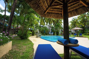 Uma área de piscina com espreguiçadeiras, um guarda-sol de palha e palmeiras.