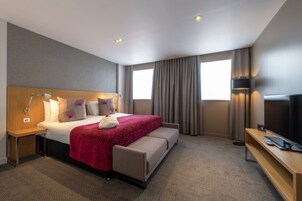 Quarto de hotel com cama, mesa de cabeceira, luminária, televisão e janela com cortinas.