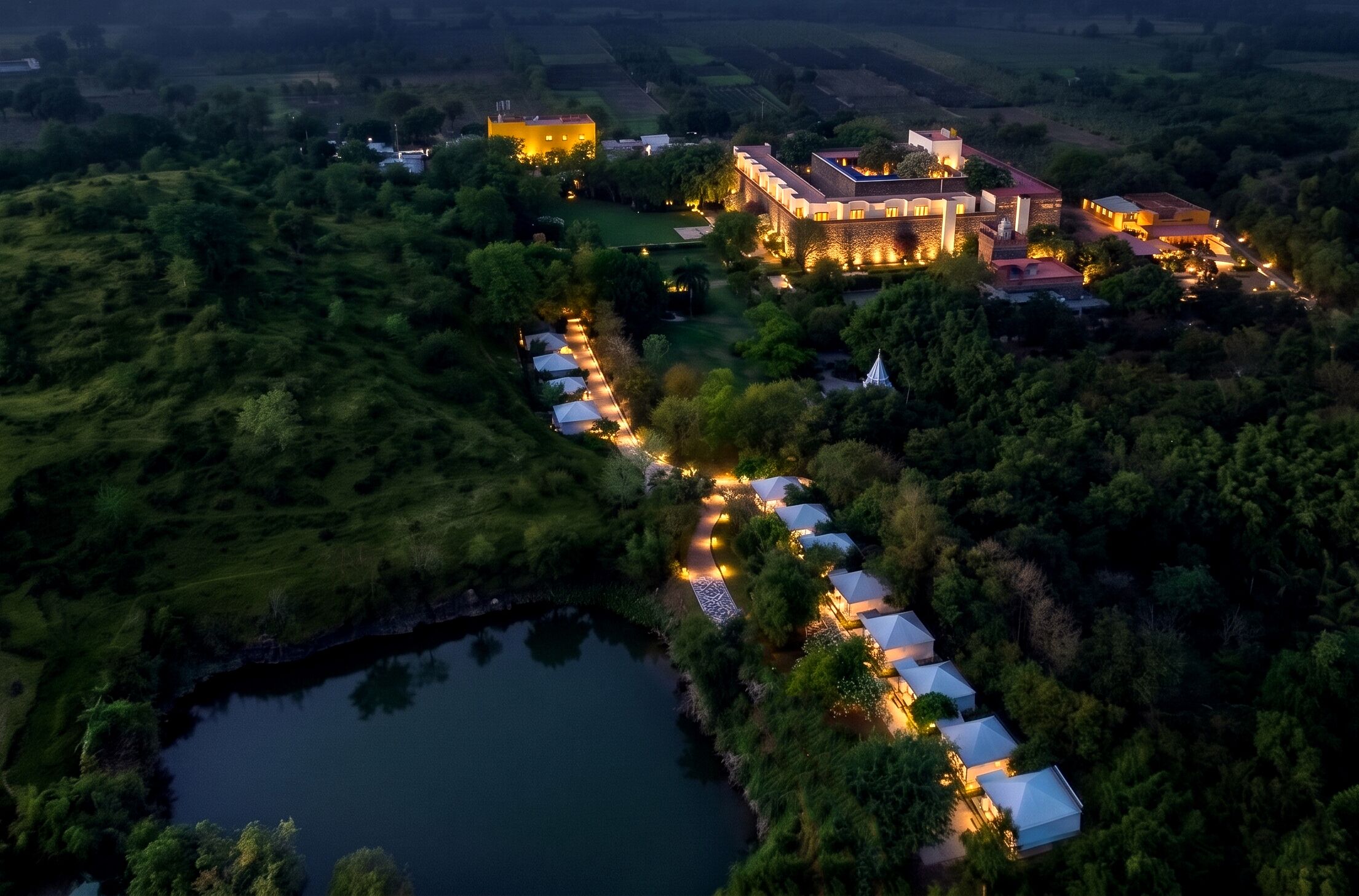 Vista aérea de um resort com um lago, edifícios interligados e um caminho bem iluminado.