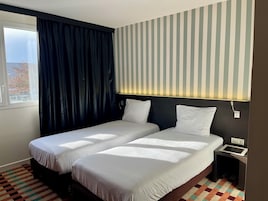 Quarto de hotel com duas camas, uma janela com cortinas, uma parede listrada e uma pequena mesa de cabeceira.