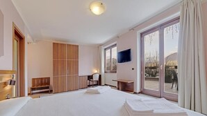 Quarto de hotel com uma cama grande, guarda-roupa de madeira, uma escrivaninha, uma cadeira, televisão e porta de vidro que desliza para o exterior.