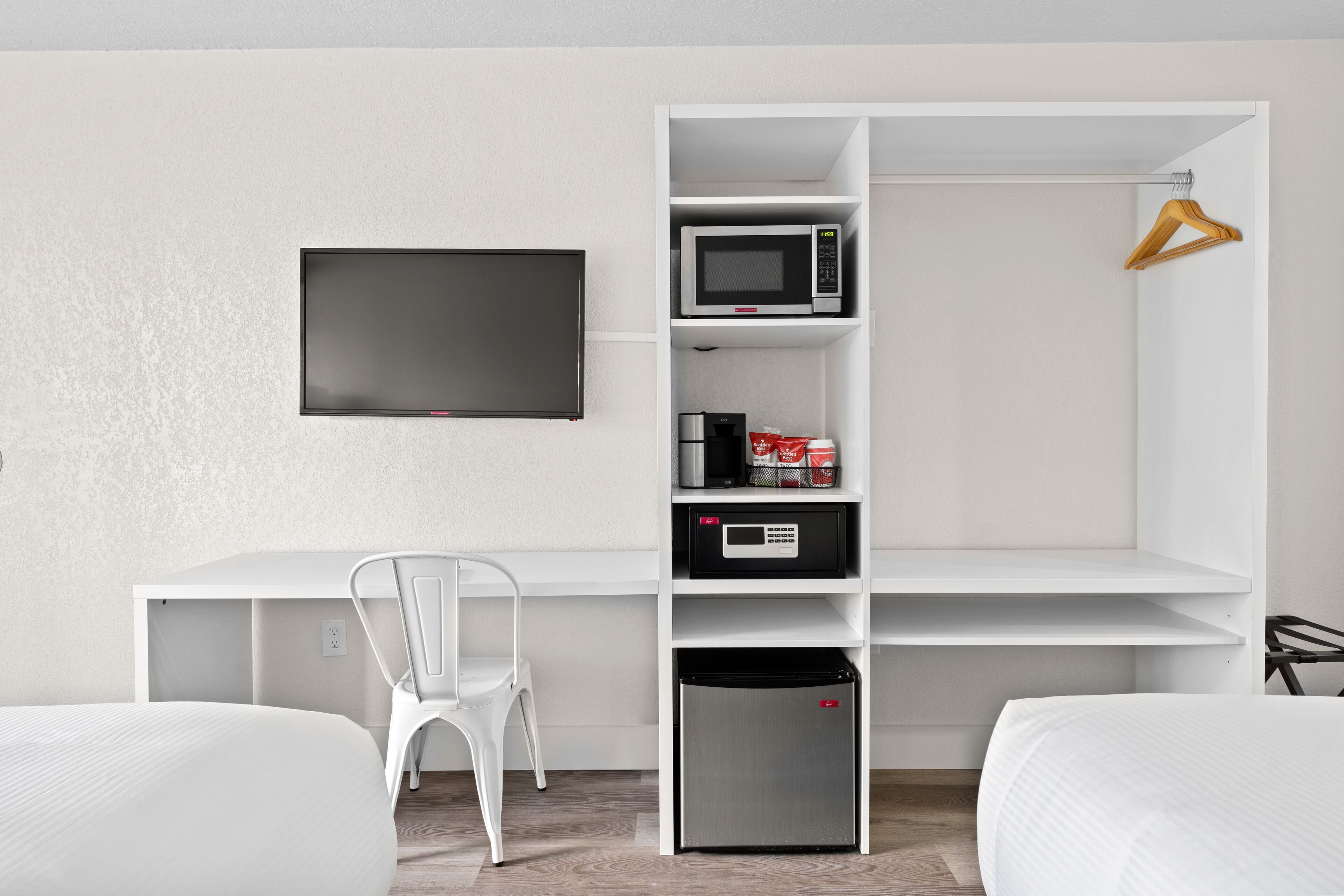 Quarto moderno com cama branca, escrivaninha branca, televisão, micro-ondas e mini-geladeira.