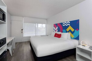 Quarto de hotel moderno com cama, escrivaninha, cadeira, televisão e janela com persianas.