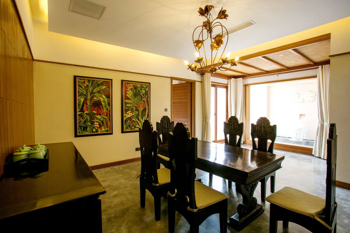 Uma sala de jantar com uma mesa de madeira grande e seis cadeiras, duas pinturas na parede e um ventilador de teto.