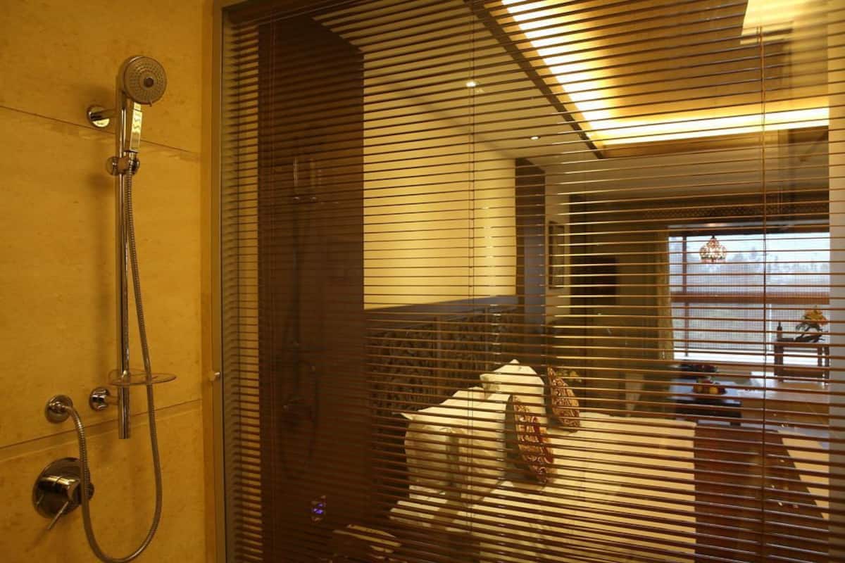 Uma casa de banho com chuveiro, sanita e lavatório. Há uma janela com persianas, e é visível um reflexo de um quarto através da janela.