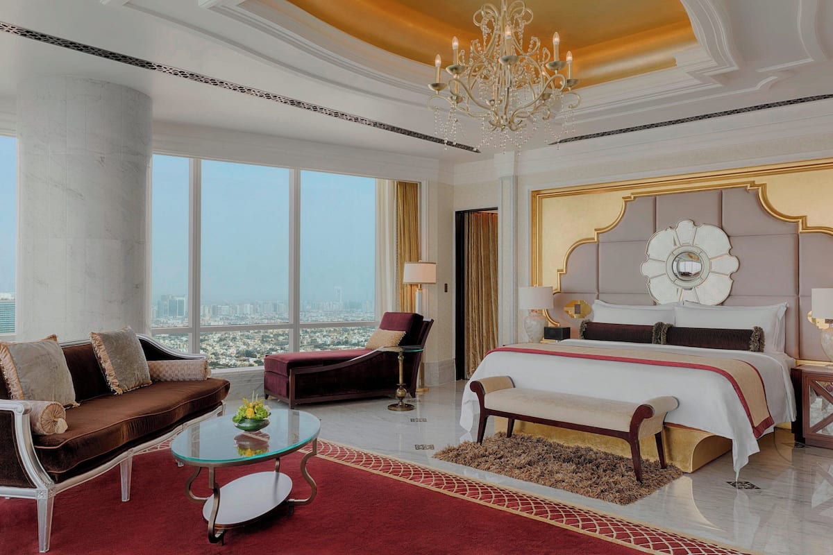 Quarto de hotel luxuoso com uma cama grande, área de estar com sofá e poltrona, mesa de centro de vidro e vista para a cidade.