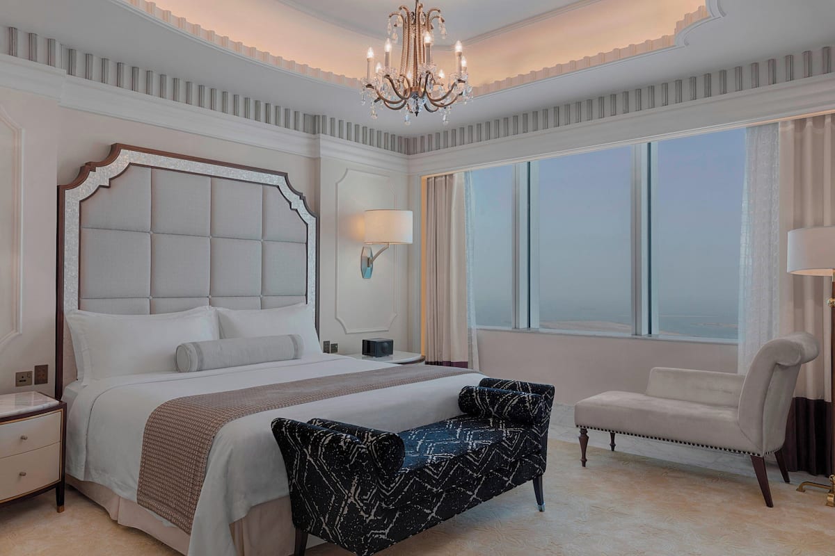 Um quarto espaçoso com uma cama grande, um lustre, uma janela com cortinas e um banco.