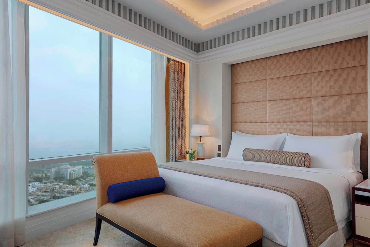 Quarto de hotel com uma grande janela, uma cama com roupa branca, um banco e uma mesa de cabeceira com abajur.