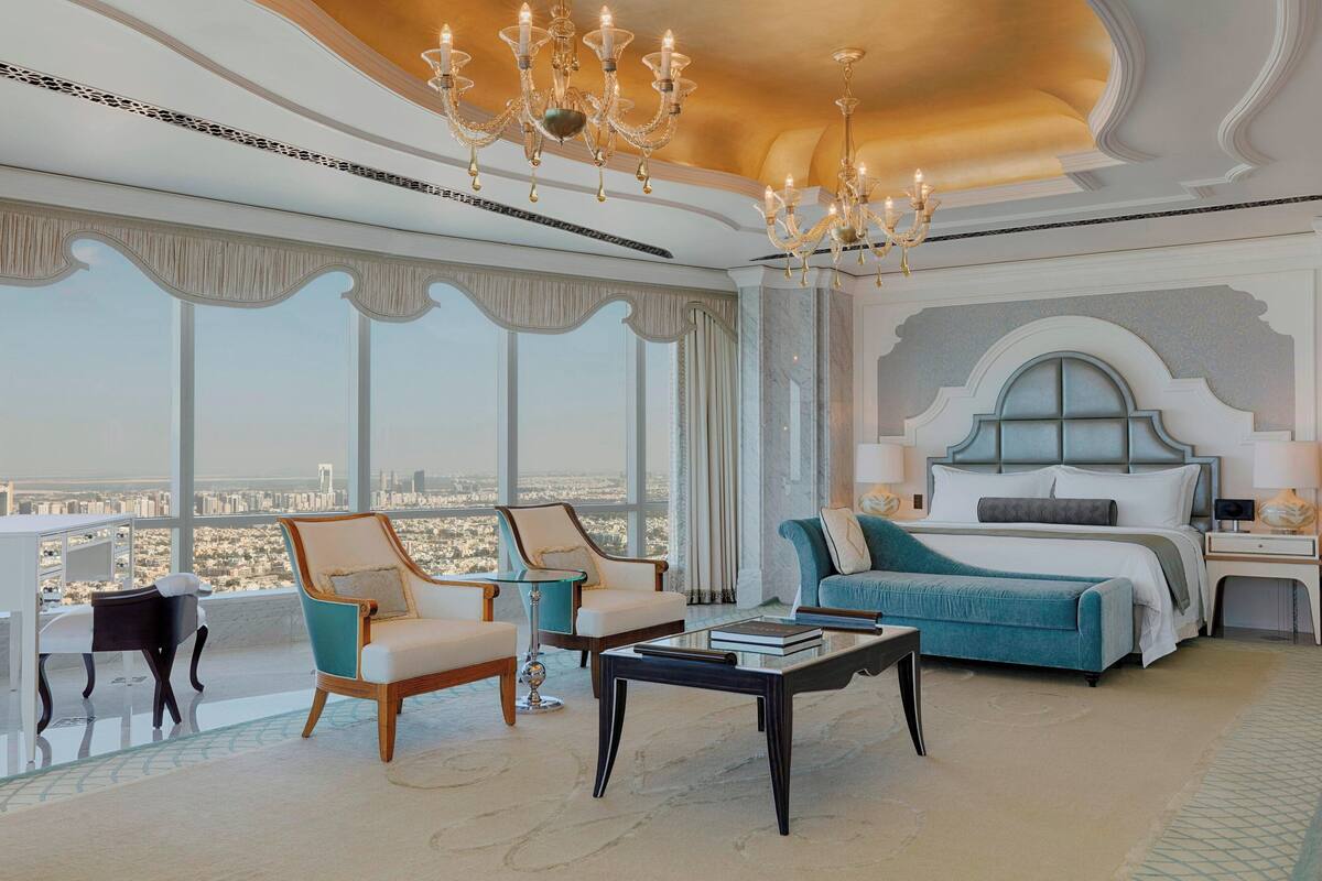Um quarto luxuoso com uma cama grande, um espaço de estar com cadeiras, uma mesa de centro e vista para um horizonte urbano através de grandes janelas.