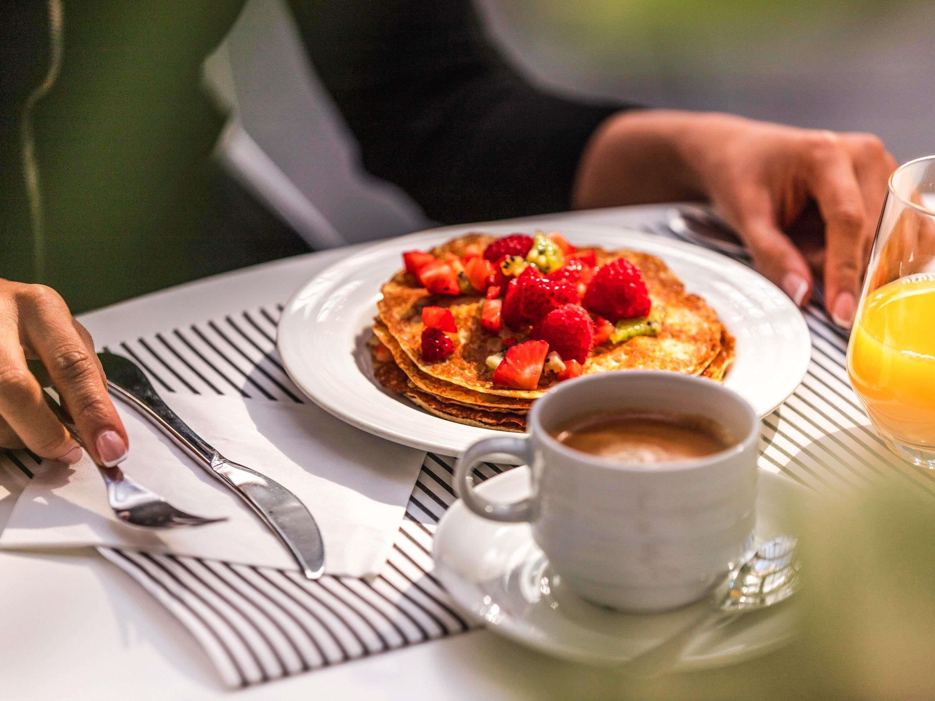 Disposição de café da manhã com panquecas cobertas de morangos e kiwi, uma xícara de café e um copo de suco de laranja sobre uma mesa.