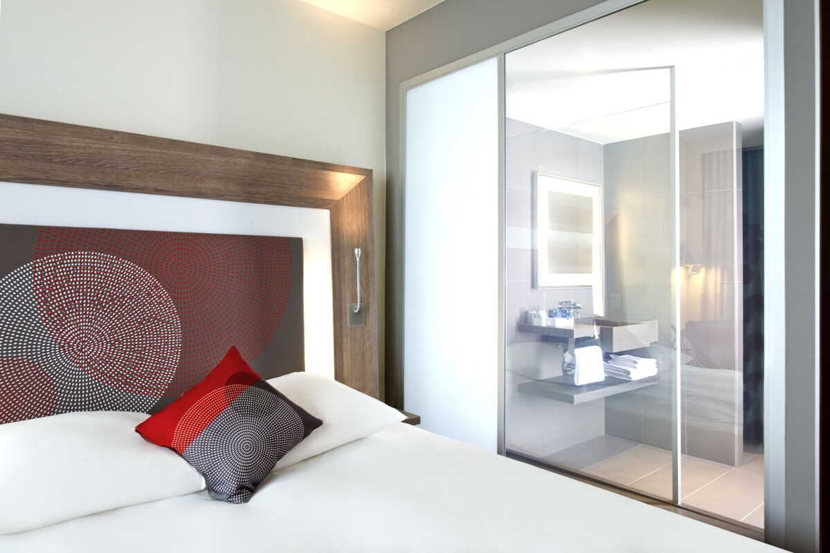Quarto de hotel moderno com uma cama grande, divisória de vidro e banheiro visível, com pia e espelho.