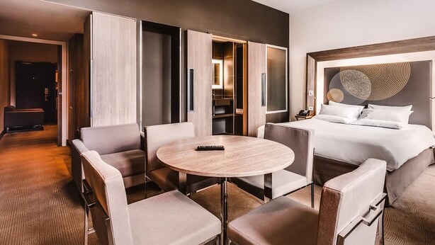 Quarto de hotel com cama, mesa redonda com cadeiras, guarda-roupa e vista para o corredor.