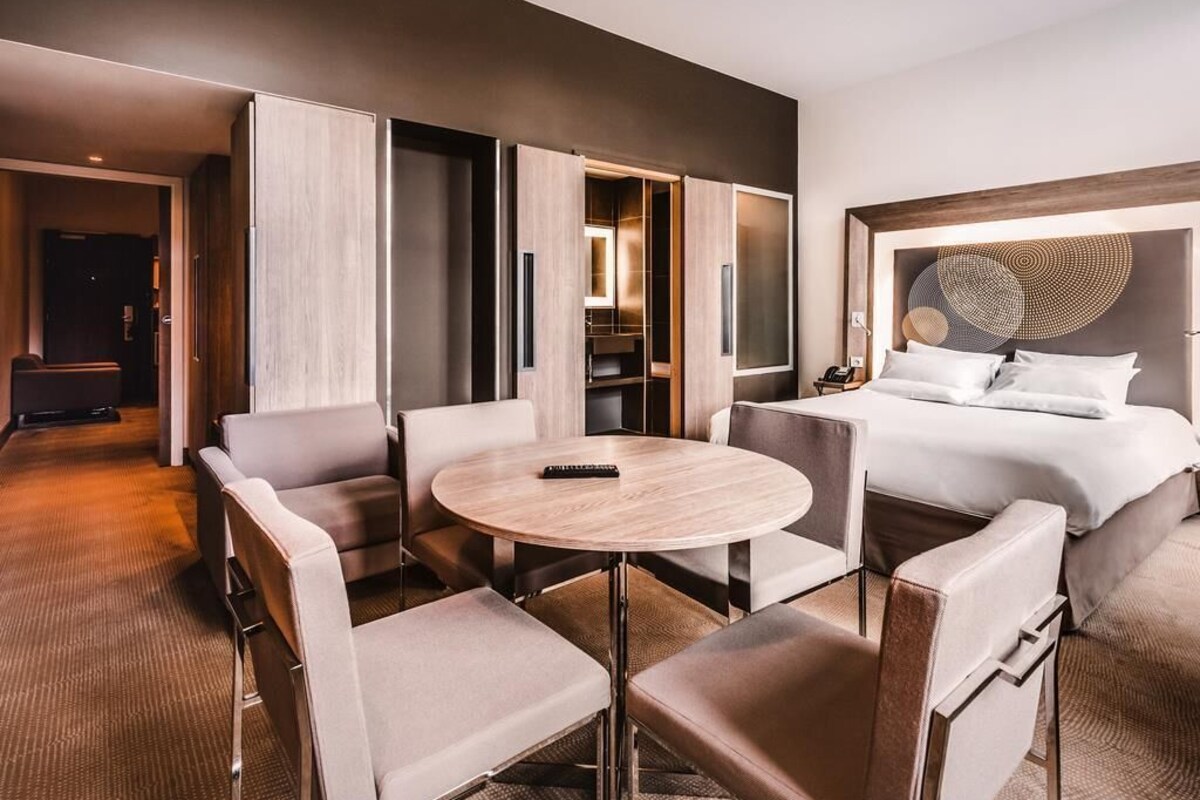 Quarto de hotel com cama, mesa redonda com cadeiras, guarda-roupa e vista para o corredor.