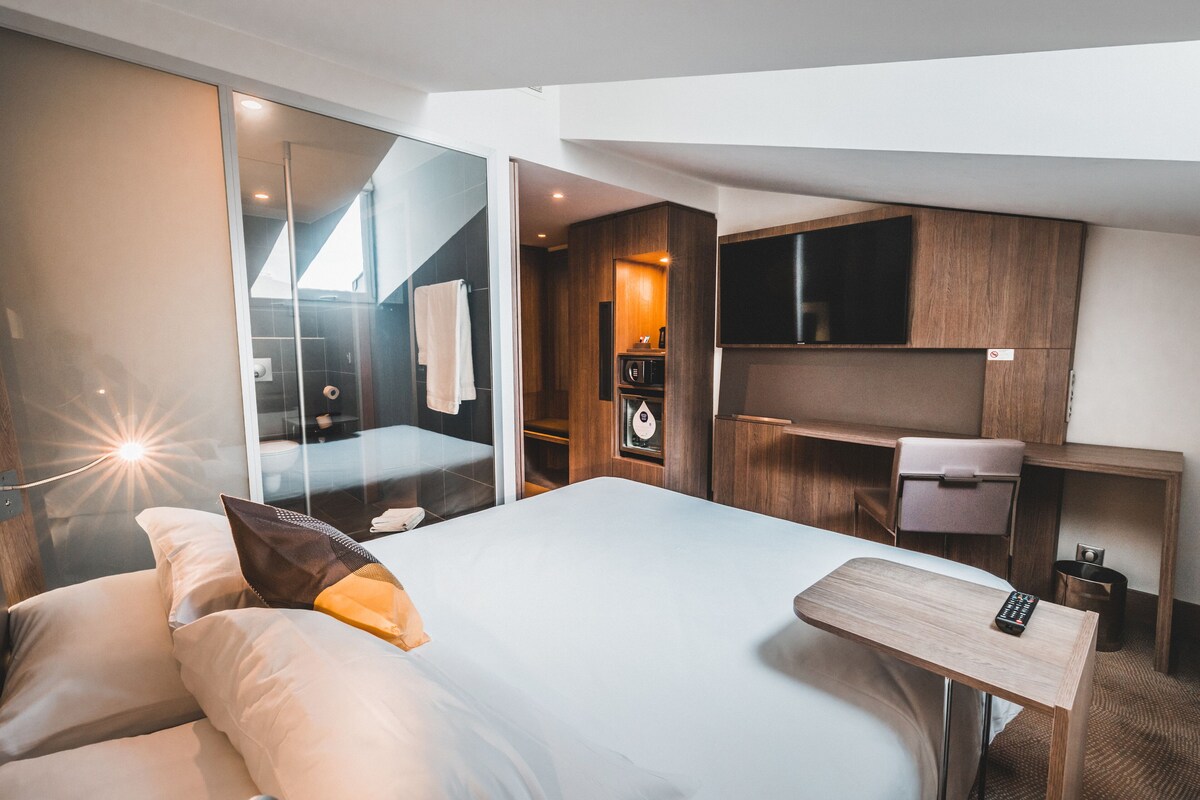 Quarto de hotel moderno com uma cama grande, uma escrivaninha, uma televisão e um banheiro visível através de um painel de vidro.