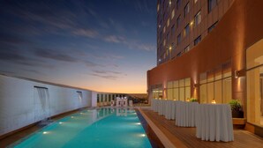 Piscina com vista para o pôr do sol, um deck de madeira e um edifício com grandes janelas.