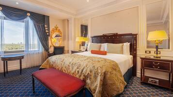 Quarto de hotel com uma cama grande, um banco, uma mesa de cabeceira, um espelho e uma janela com cortinas.