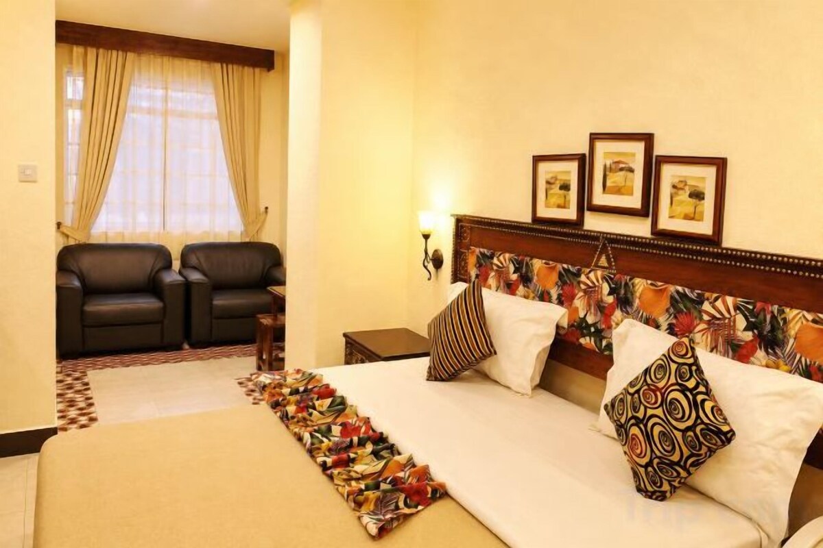 Quarto de hotel com uma cama, duas cadeiras, uma mesa de cabeceira e uma luminária fixada na parede.