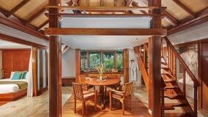 Um quarto espaçoso com uma mesa de jantar de madeira rodeada de cadeiras, uma janela grande com vista para o verde e uma escada de madeira.