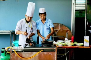 Dois chefs cozinhando em uma cozinha com fogão, panelas e uma mesa com pratos e temperos.
