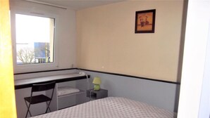 Quarto pequeno e compacto com uma cama de solteiro, uma escrivaninha com cadeira, uma mesinha de cabeceira com abajur e uma janela.