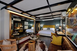 Quarto de hotel com uma cama grande, área de estar com sofá de couro, mesa de jantar e um espelho refletindo o ambiente.