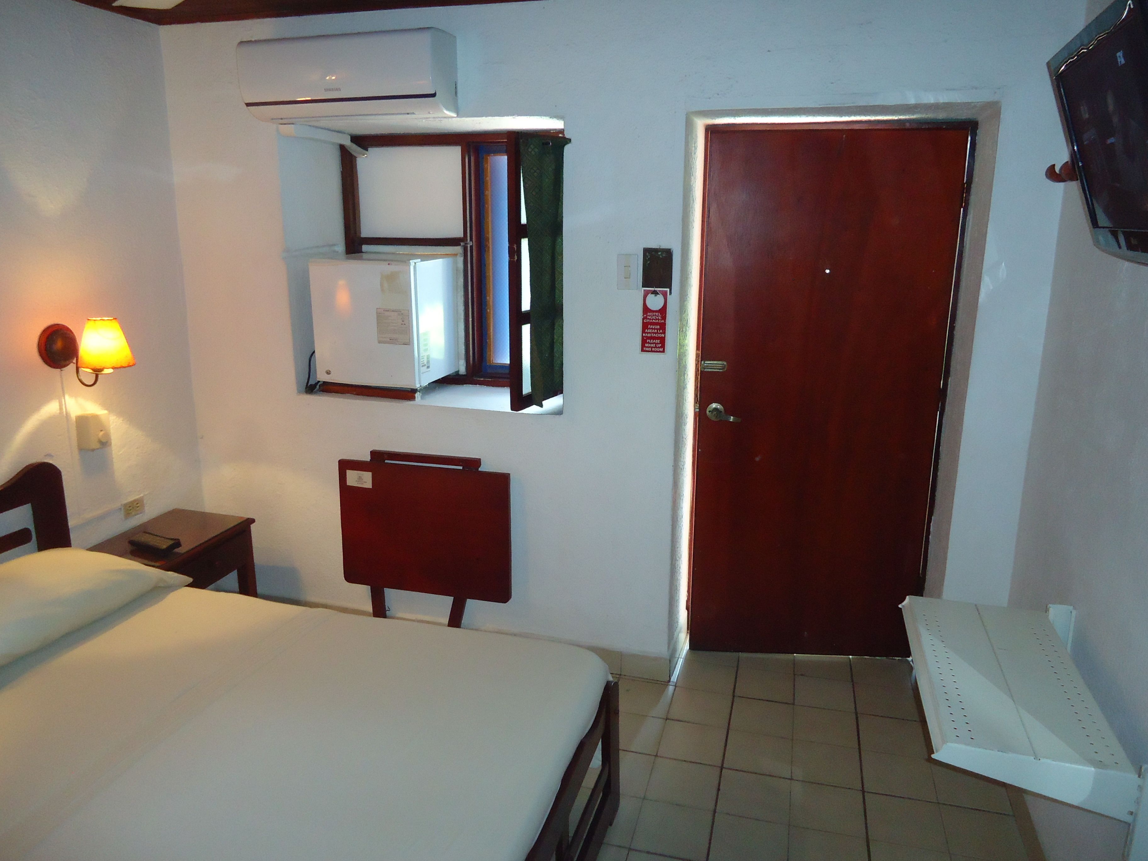 Quarto de hotel com cama, escrivaninha com luminária, espelho, geladeira e porta com placa.