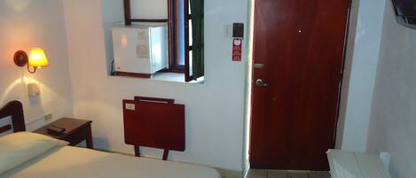 Quarto de hotel com cama, escrivaninha com luminária, espelho, geladeira e porta com placa.