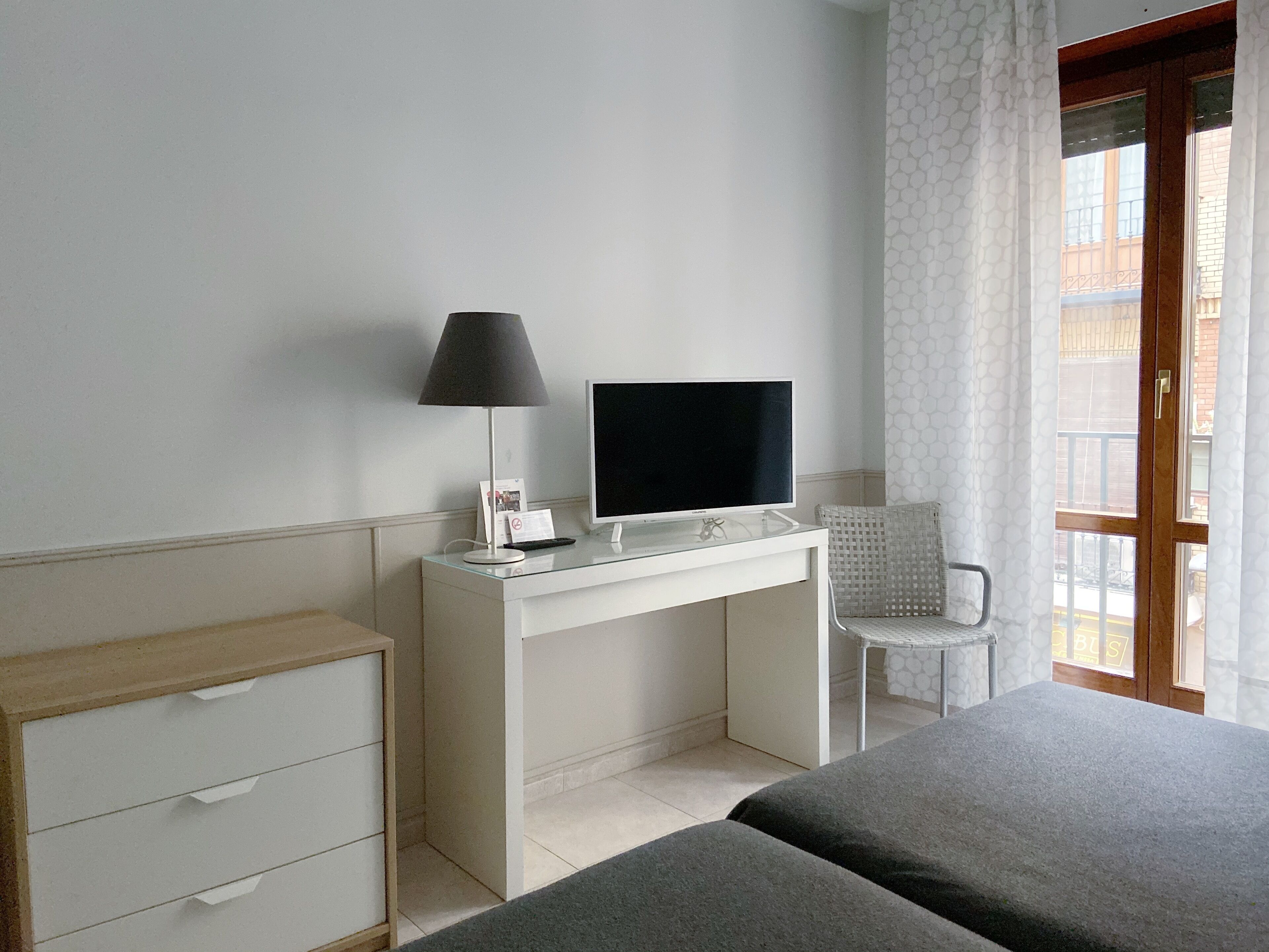 Quarto de hotel com secretária branca, monitor de computador, luminária, cadeira, cama e janela com vista para o exterior.