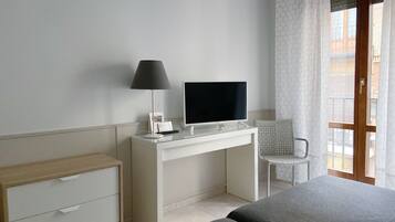 Quarto de hotel com secretária branca, monitor de computador, luminária, cadeira, cama e janela com vista para o exterior.