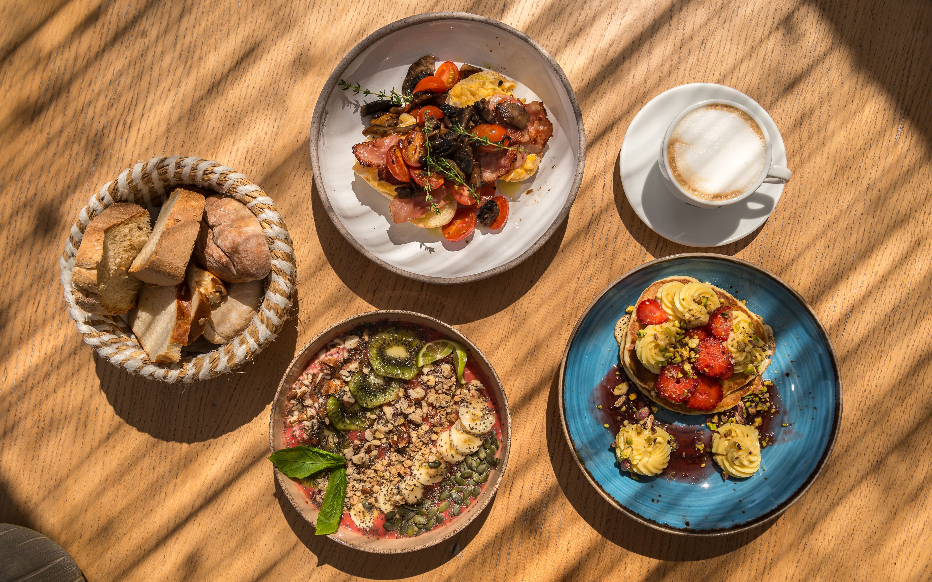 Uma mesa com quatro pratos: uma cesta de pão, um prato com legumes e carne, uma tigela com mistura de frutas e nozes, e um prato com panquecas e morangos.