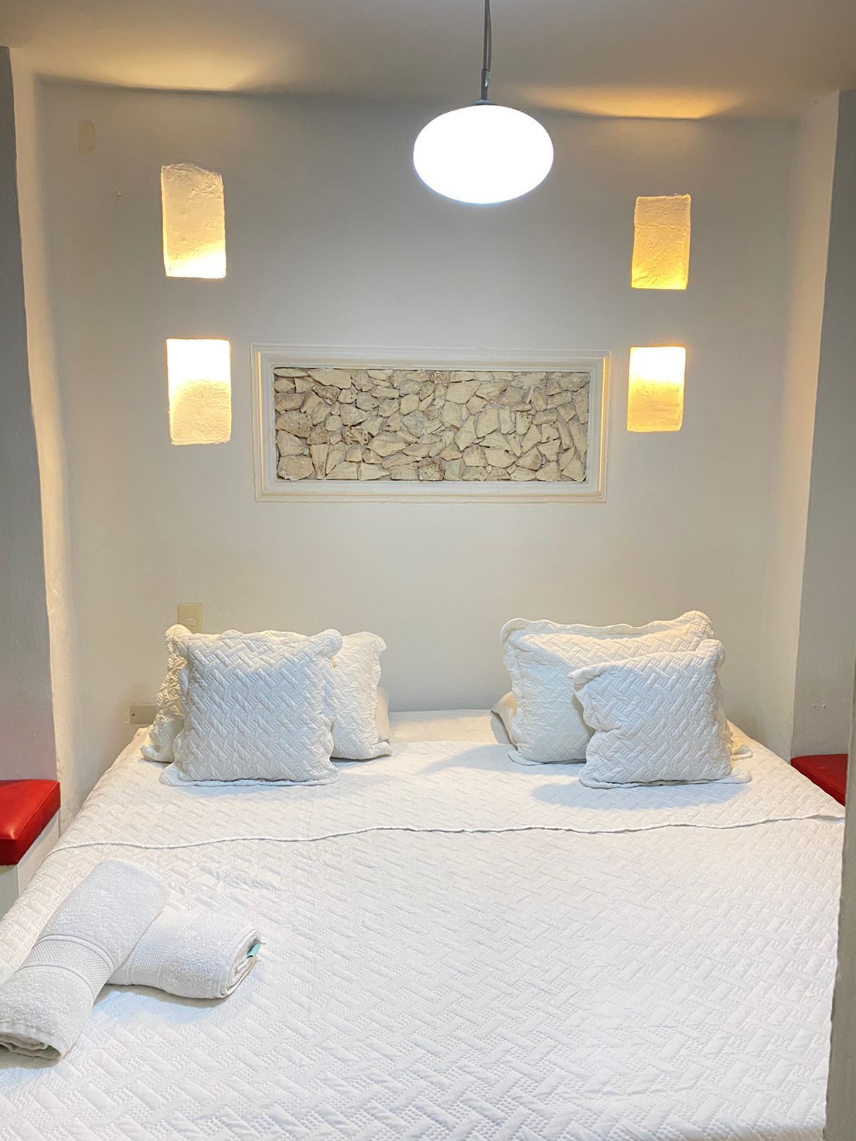 Cama bem arrumada com dois travesseiros e uma toalha enrolada, uma obra de arte abstrata na parede e duas luminárias de parede.