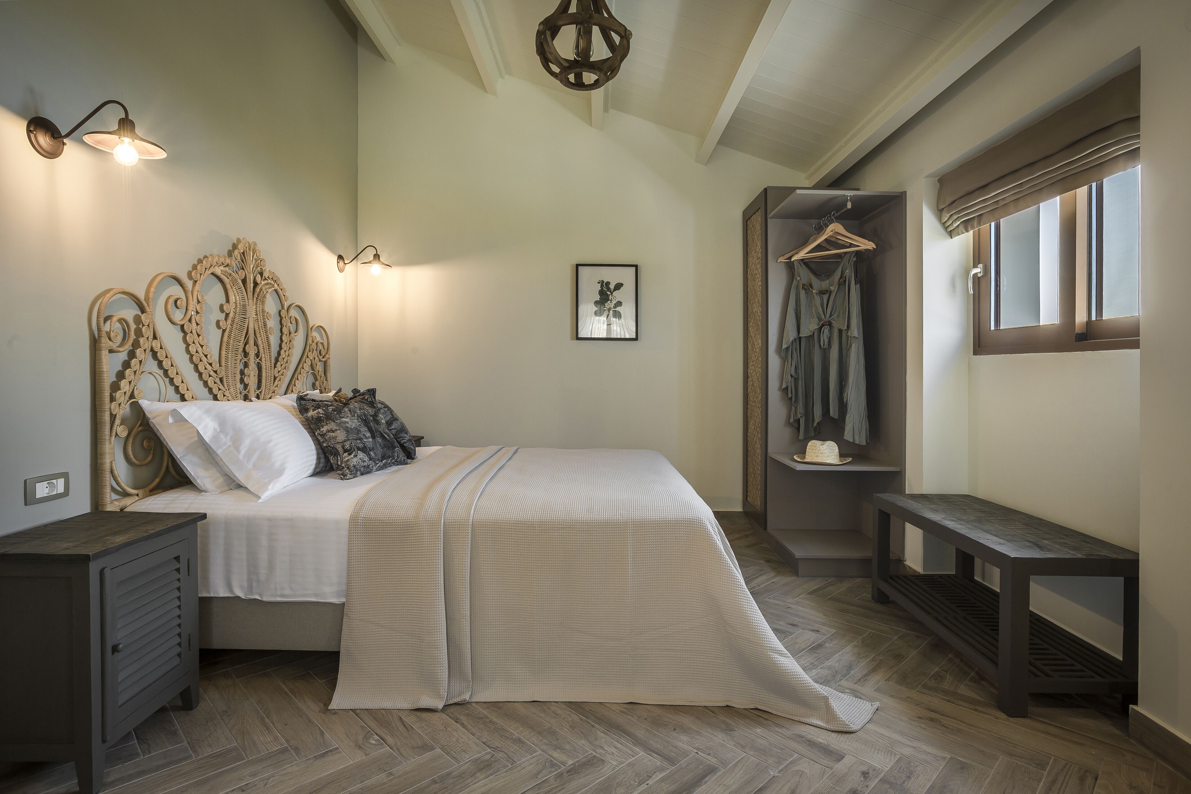 Quarto com uma cama grande, cabeceira de madeira, mesa de cabeceira, um banquinho, luminária pendente, quadro emoldurado e janela com cortinas.