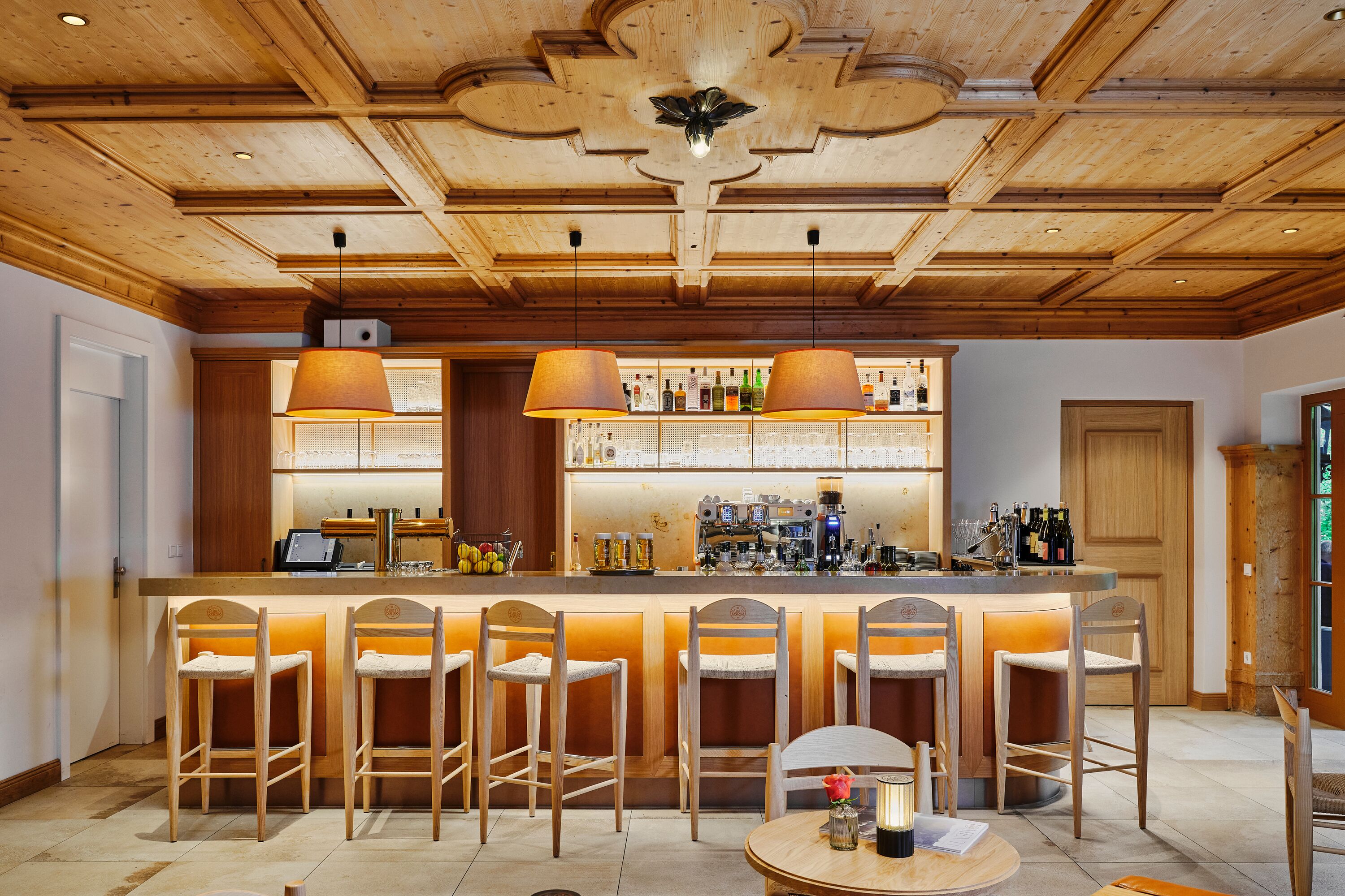 Uma área de bar com teto de madeira, banquetas e um balcão com diversos frascos e copos.
