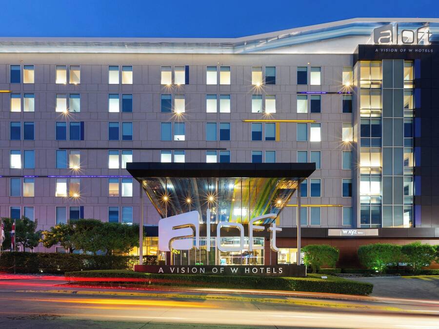 Um edifício de hotel moderno com uma placa chamativa 'aloft', vários andares e uma entrada bem iluminada.