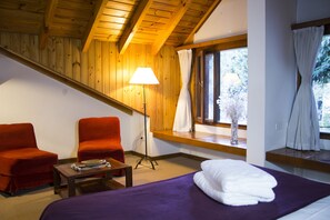 Um quarto com uma cama, duas cadeiras vermelhas, uma mesinha, um abajur e uma janela com cortinas.