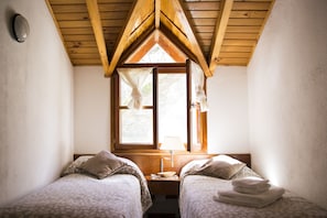 Quarto com duas camas, janela com cortinas, teto de madeira e mesa de cabeceira com abajur.
