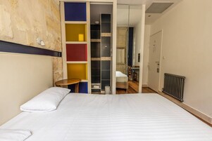 Quarto de hotel com cama, mesa redonda pequena, guarda-roupa com painéis coloridos, radiador e banheiro visível por uma porta aberta.