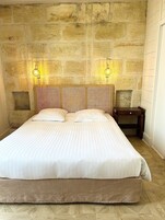 Uma cama com cabeceira de madeira e duas luminárias fixadas na parede, diante de uma parede de pedra.