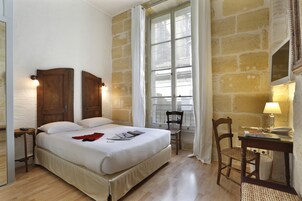Quarto com cama grande, cabeceira de madeira, uma cadeira, escrivaninha com abajur e janela com cortinas.